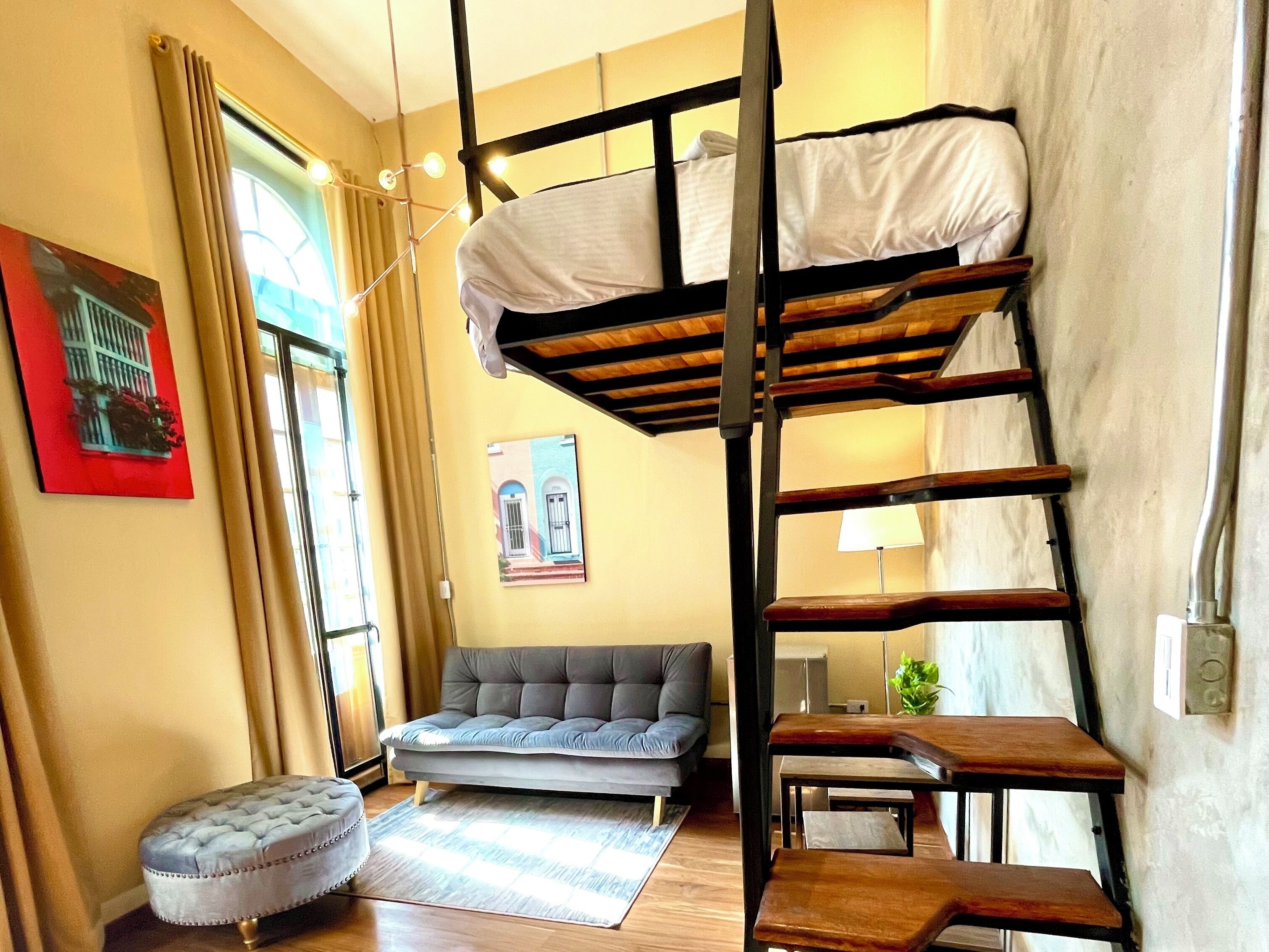 Quarto compacto com cama beliche, sofá, um pequeno banquinho e uma luminária.