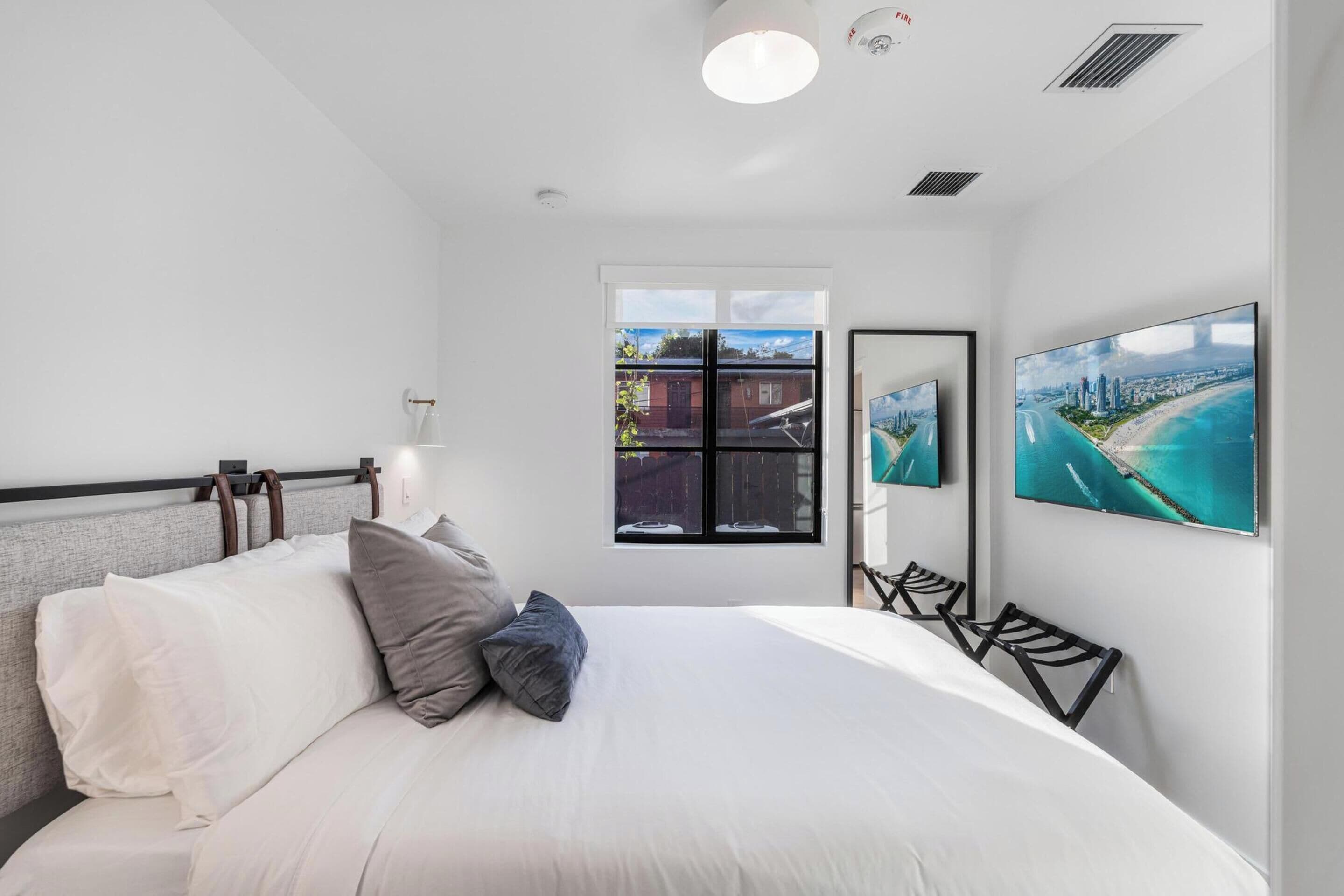 Quarto de hotel moderno com uma cama grande, uma janela vista para a cidade, um quadro de uma praia e uma luminária no teto.