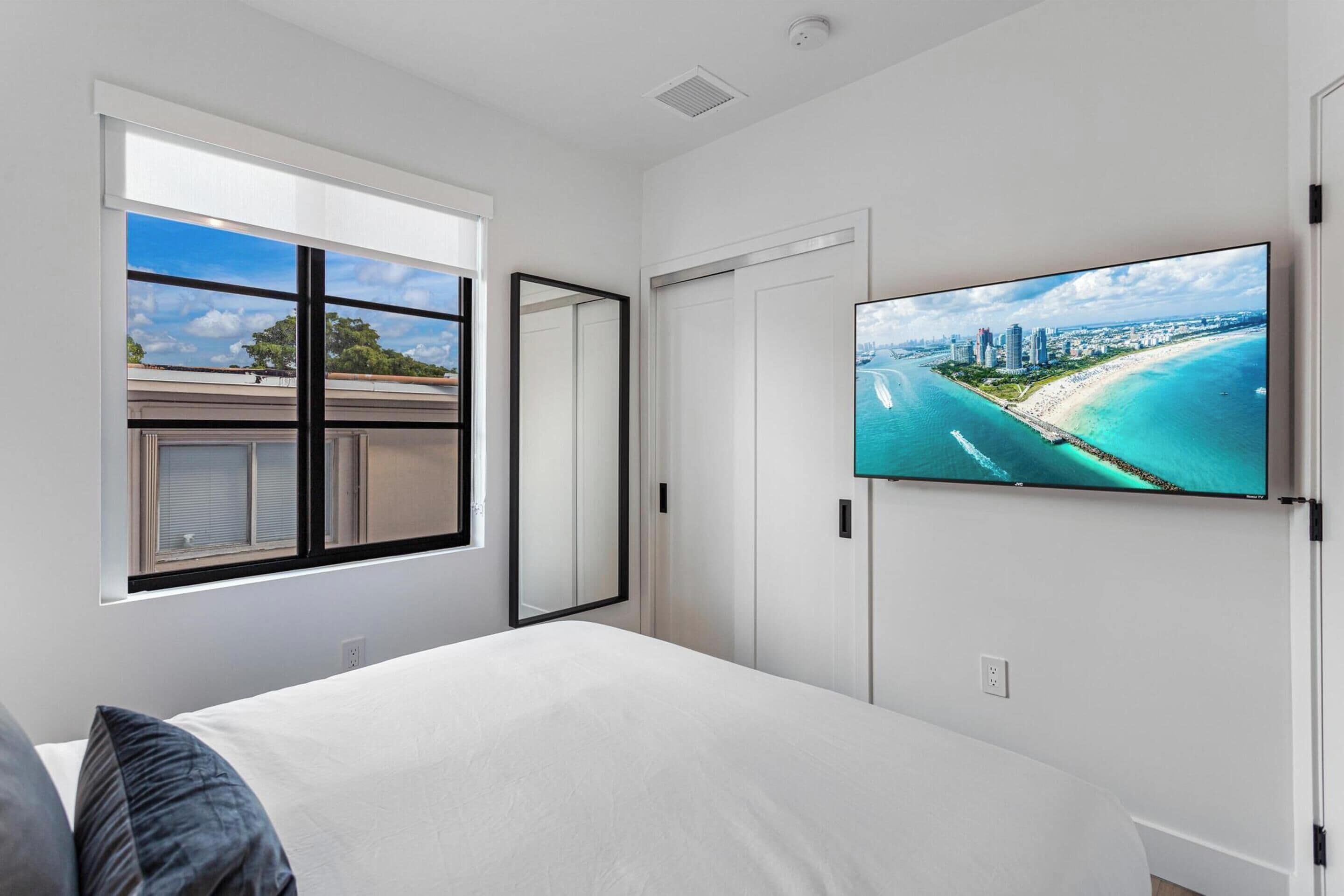 Quarto com uma janela grande, uma cama, uma televisão na parede e uma porta.