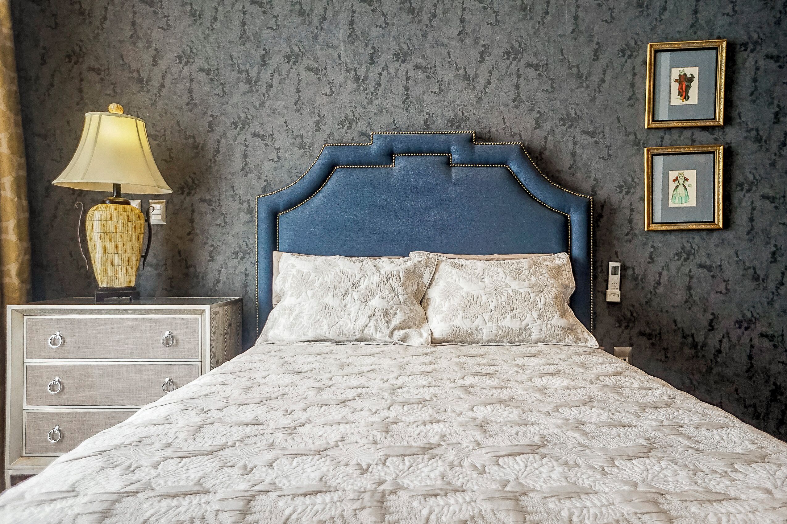 Quarto com cabeceira estofada azul, colcha branca acolchoada, mesa de cabeceira com abajur e duas molduras na parede.