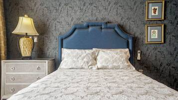 Quarto com cabeceira estofada azul, colcha branca acolchoada, mesa de cabeceira com abajur e duas molduras na parede.