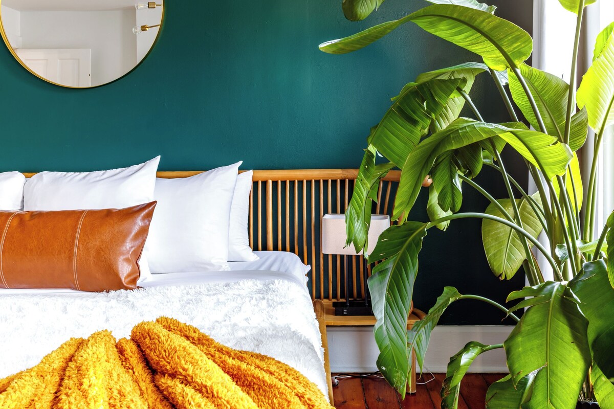 Quarto com cama, espelho redondo, planta grande e janela.