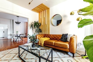 Uma sala de estar com um sofá de couro marrom, uma mesa de centro de vidro, um tapete estampado, um painel de parede de madeira e um espelho redondo.