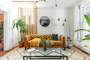 Uma sala de estar moderna com um sofá de couro marrom, uma mesa de centro de vidro, um espelho redondo, um abajur de chão e um vaso com planta.