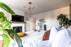 Quarto moderno com cama, roupa de cama branca, televisão sobre lareira e plantas em vasos.