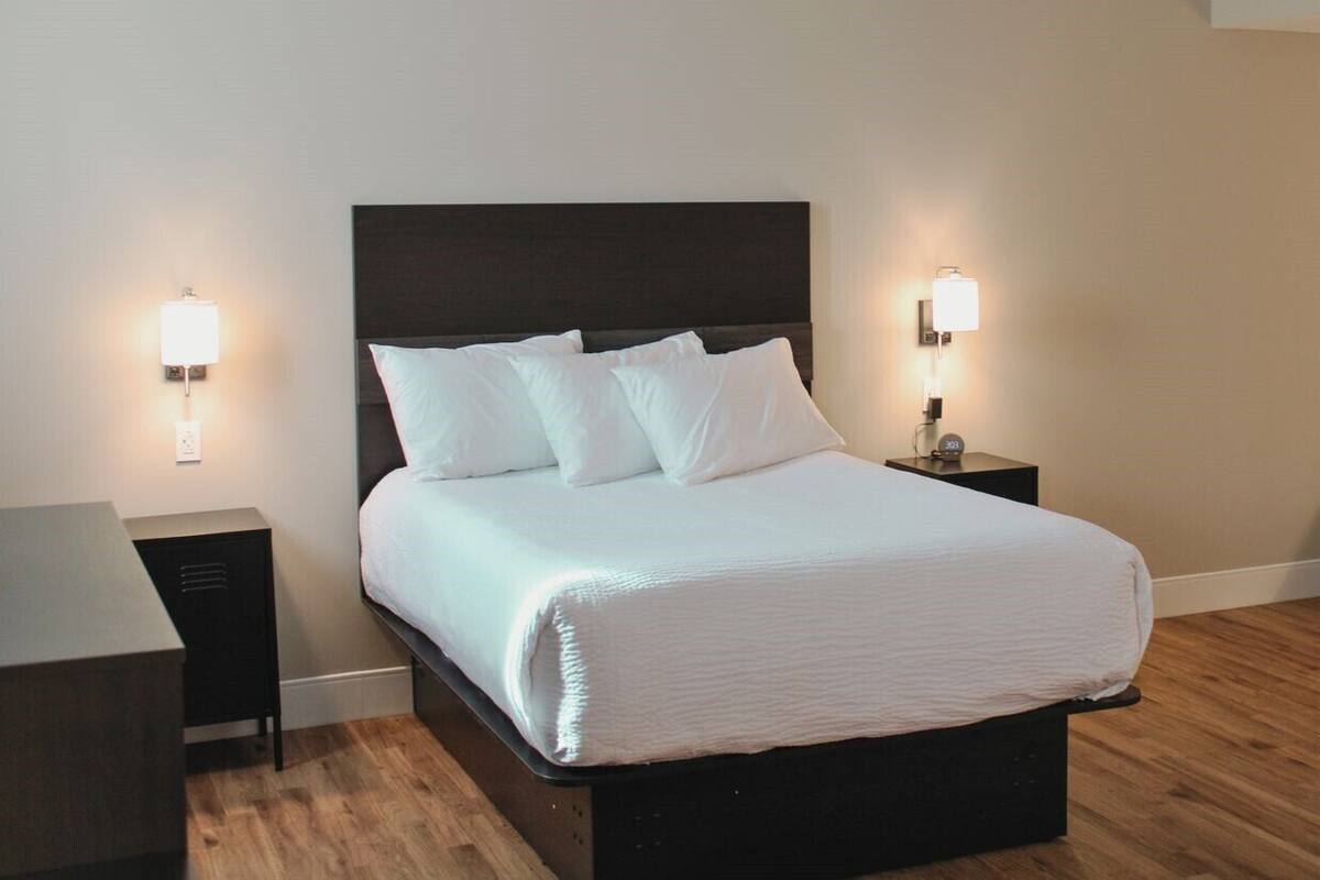 Cama bem arrumada com lençóis e almofadas brancas, acompanhada por duas mesas de cabeceira com abajures, em um quarto com piso de madeira e paredes lisas.