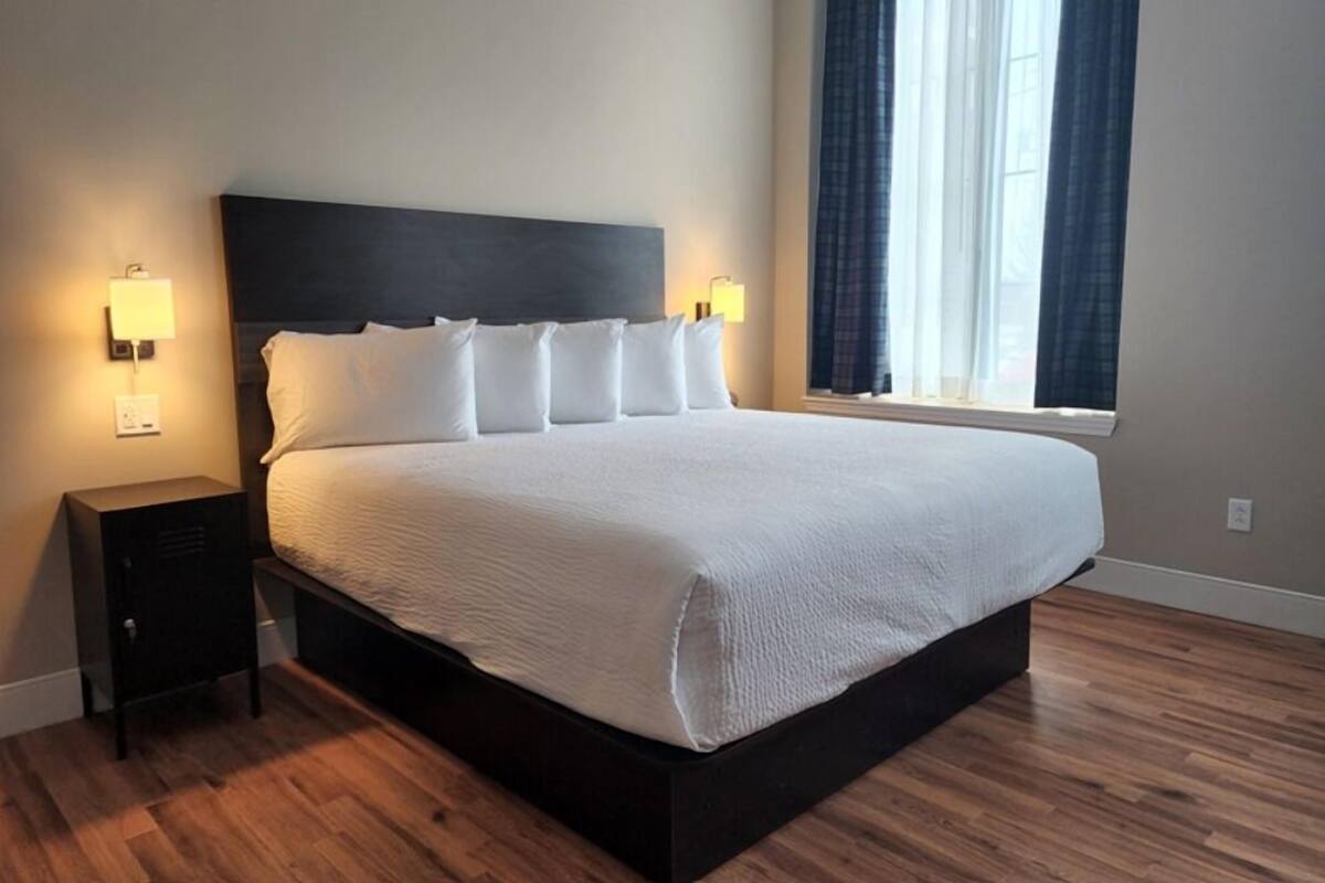Uma cama grande com cabeceira escura, duas luminárias de apoio e uma janela com cortinas.