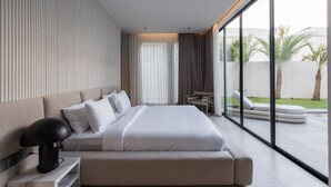 Quarto moderno com uma cama grande, uma mesinha de cabeceira com abajur, vista para o exterior através de amplas janelas e um espaço de estar com sofá e cadeiras.