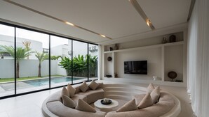 Uma sala de estar moderna com um sofá circular, uma televisão de tela plana e uma grande janela com vista para uma piscina e vegetação.