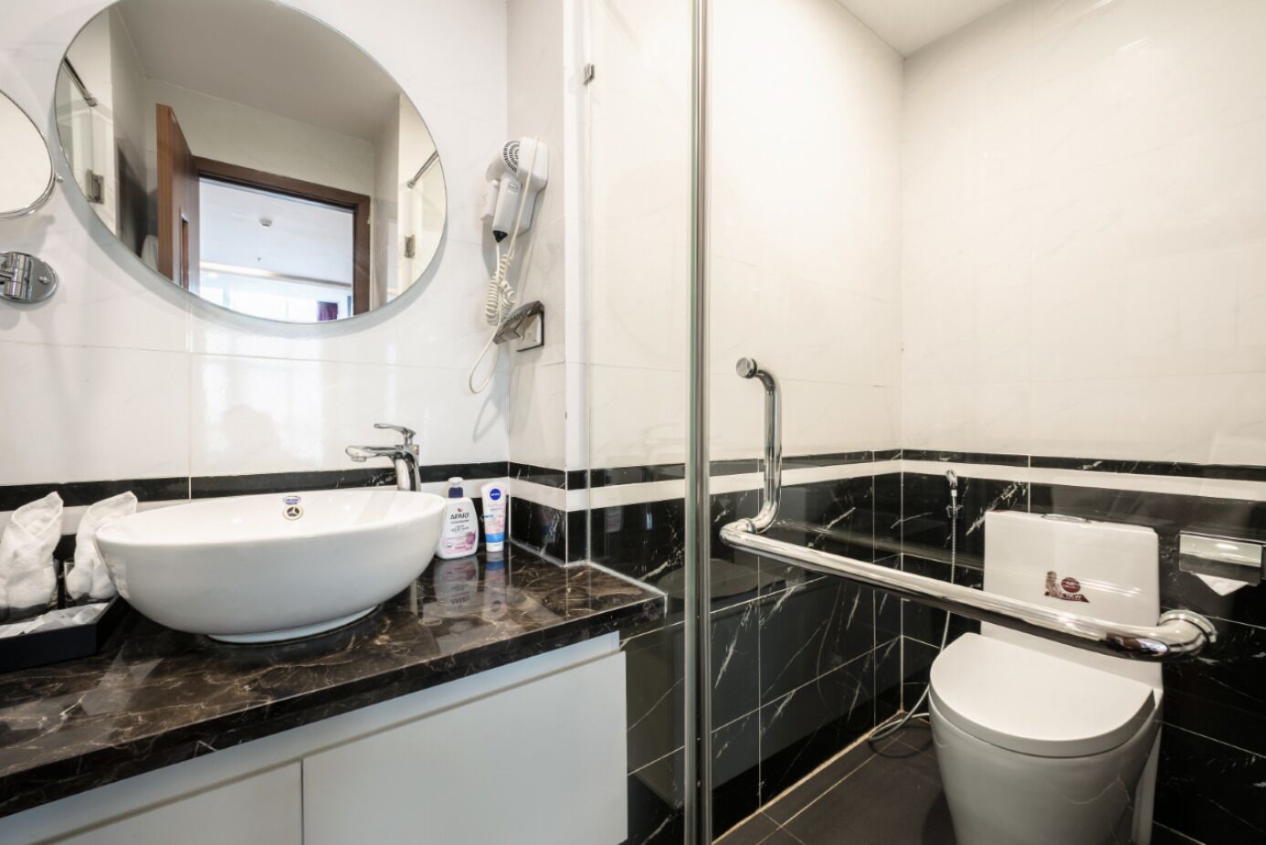 Uma casa de banho moderna com uma sanita, um lavatório branco, um espelho redondo, um secador de cabelo e uma cabine de duche em vidro.