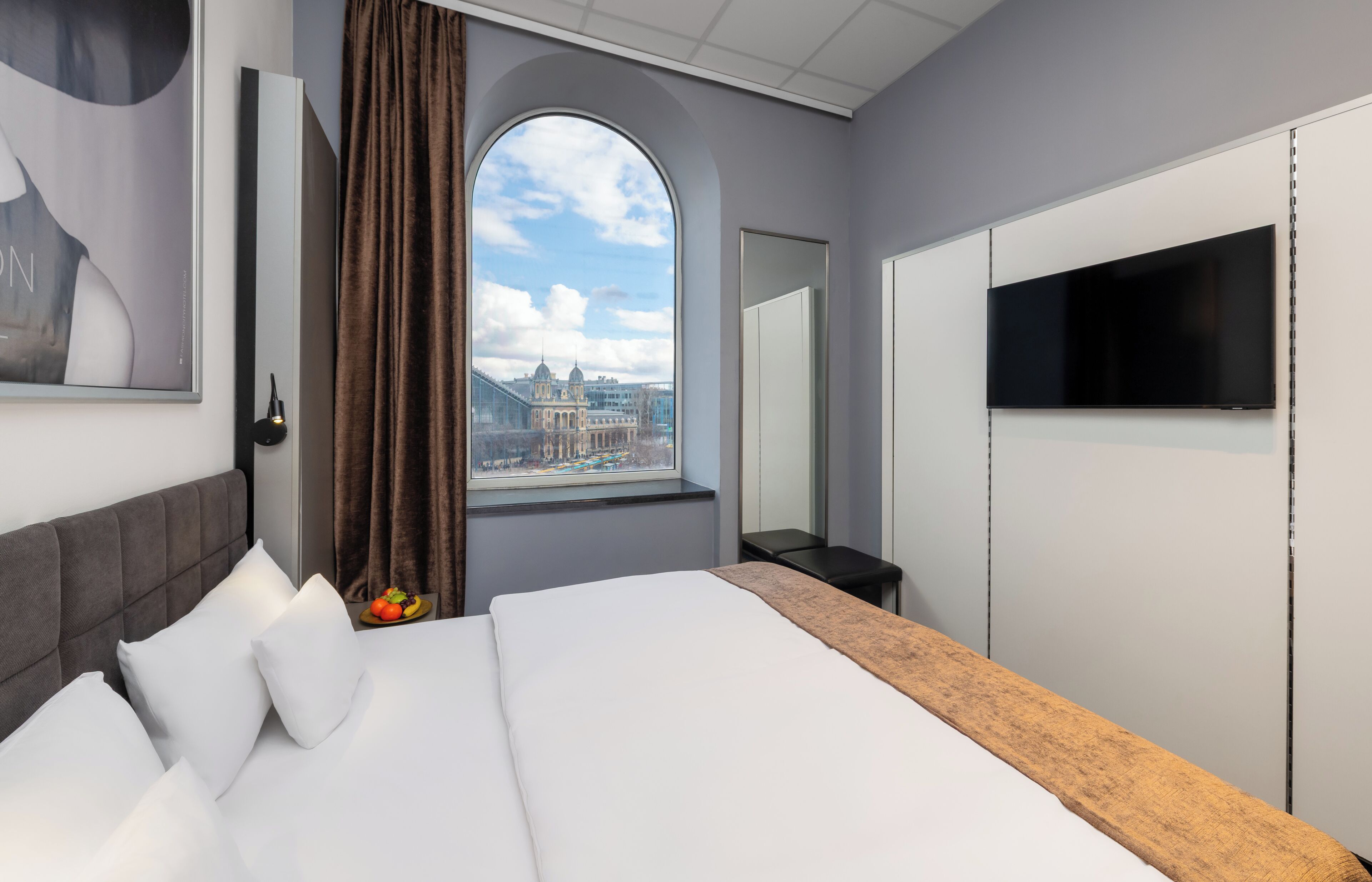 Quarto de hotel moderno com uma cama grande, televisão, guarda-roupa e janela com vista para a cidade.
