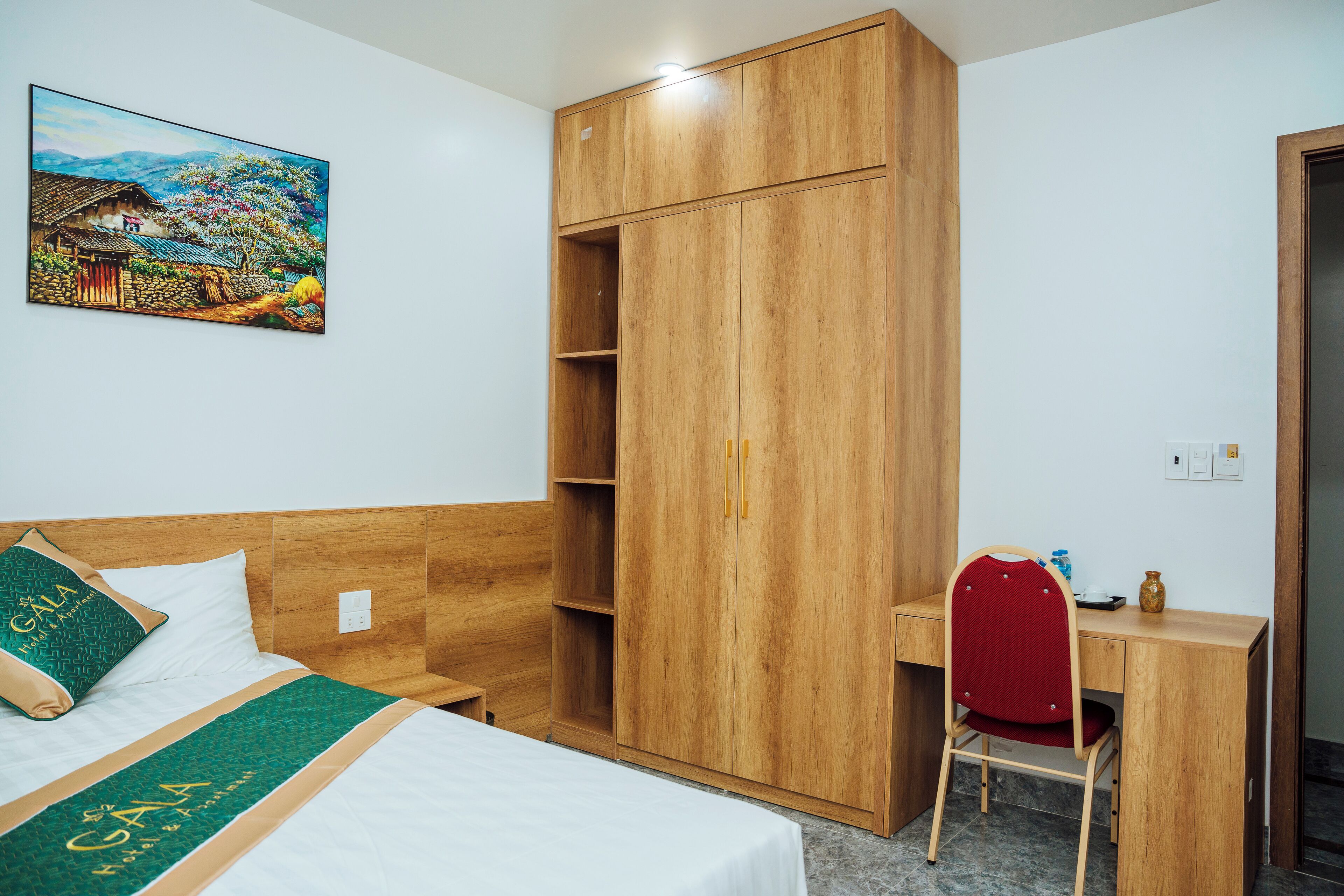 Quarto de hotel com cama, guarda-roupa de madeira, secretária com cadeira e um quadro na parede.