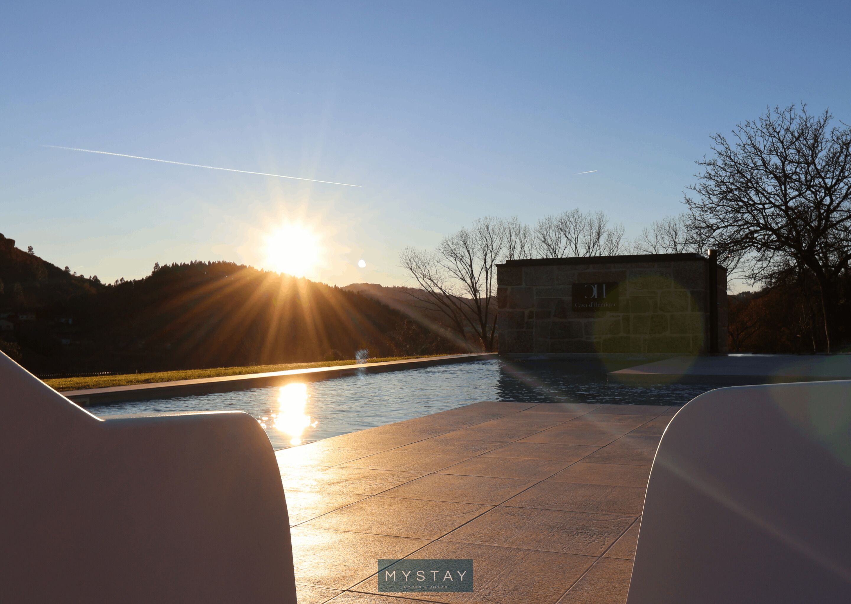 Área da piscina com um edifício de pedra com a inscrição 'Casa d'Heritage' e vista para o pôr do sol sobre a água.