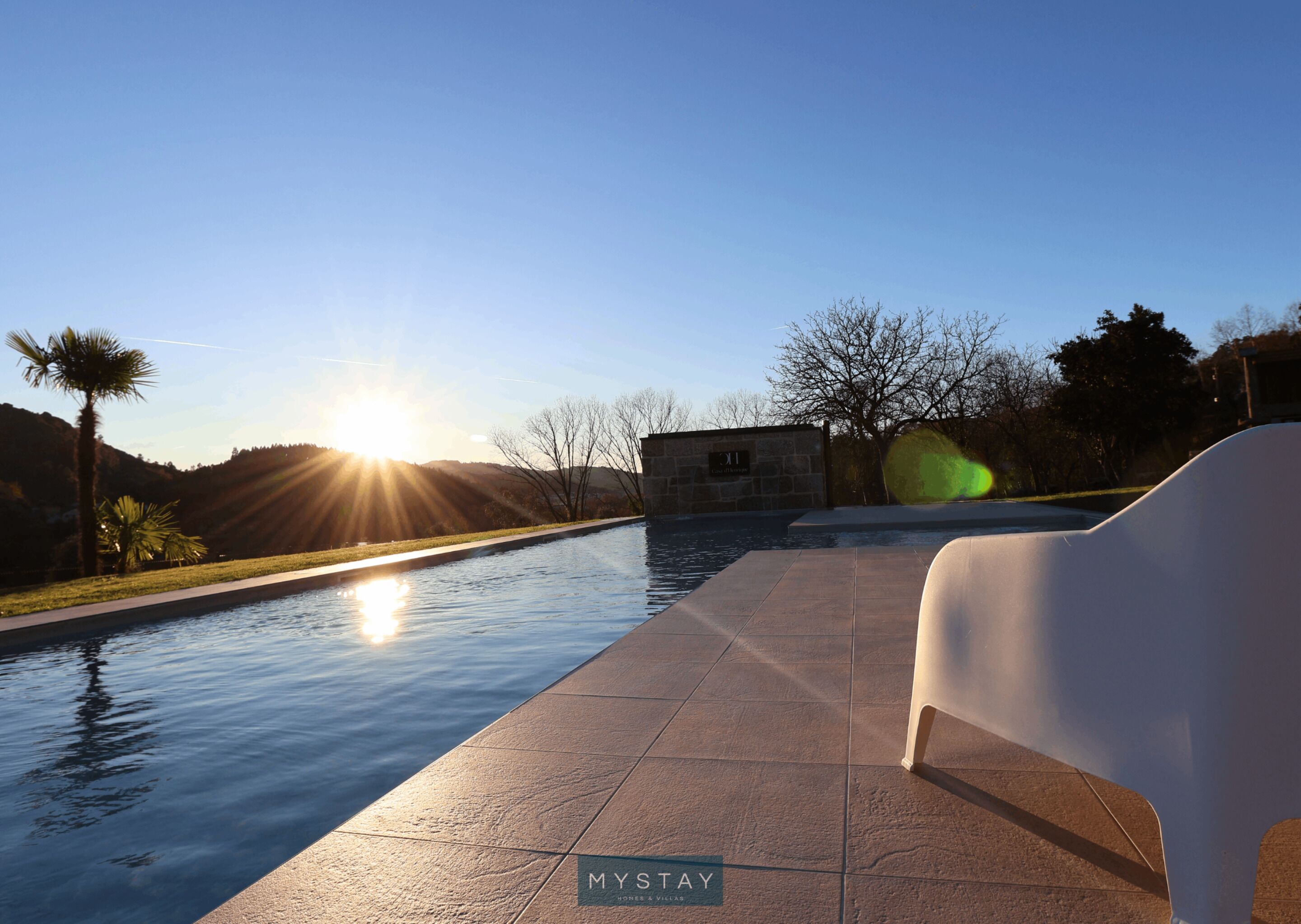 Piscina com vista desimpedida para o pôr do sol, uma parede de pedra e uma palmeira.