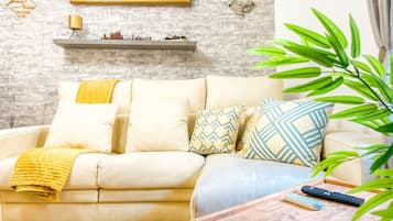 Uma sala de estar com um sofá, almofadas, uma mesa de centro, uma planta e um quadro na parede.