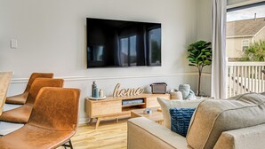 Uma sala de estar moderna com uma TV de tela plana na parede, uma mesa de console de madeira com objetos decorativos e uma janela grande com vista para uma casa.