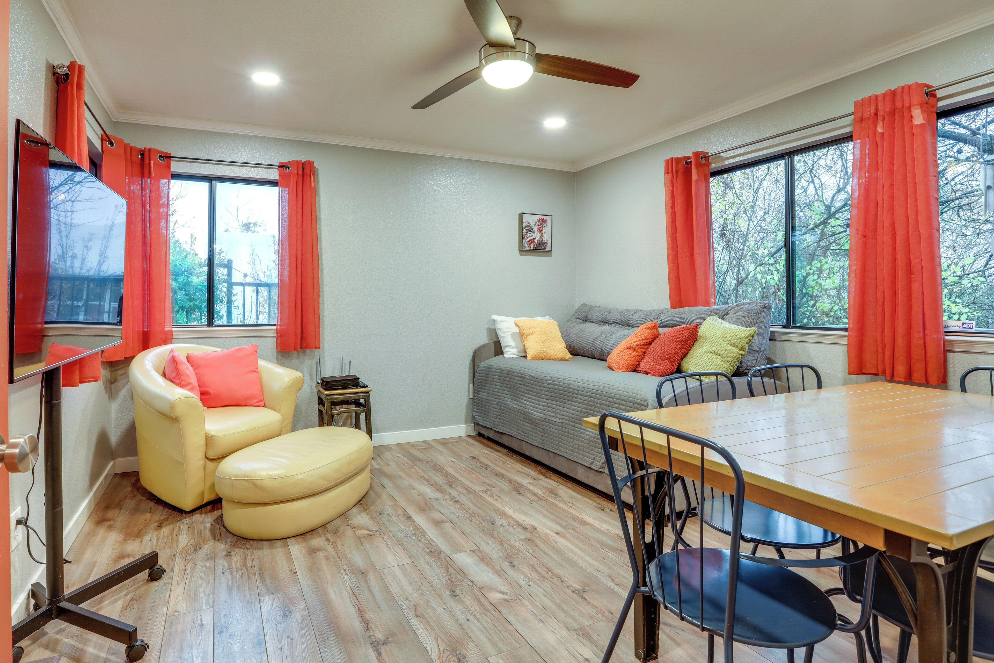 Quarto com mesa de jantar, cadeiras, sofá com almofadas, ventilador de teto e janelas com cortinas.