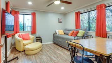 Quarto com mesa de jantar, cadeiras, sofá com almofadas, ventilador de teto e janelas com cortinas.