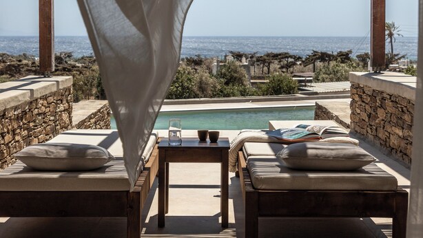 Uma área externa com mesas e cadeiras iluminada pelo sol, com piscina e vista para o mar.