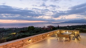 Um pátio ao ar livre com paredes de pedra e uma mesa com cadeiras, com vista para uma paisagem costeira com edifícios e montanhas ao longe.