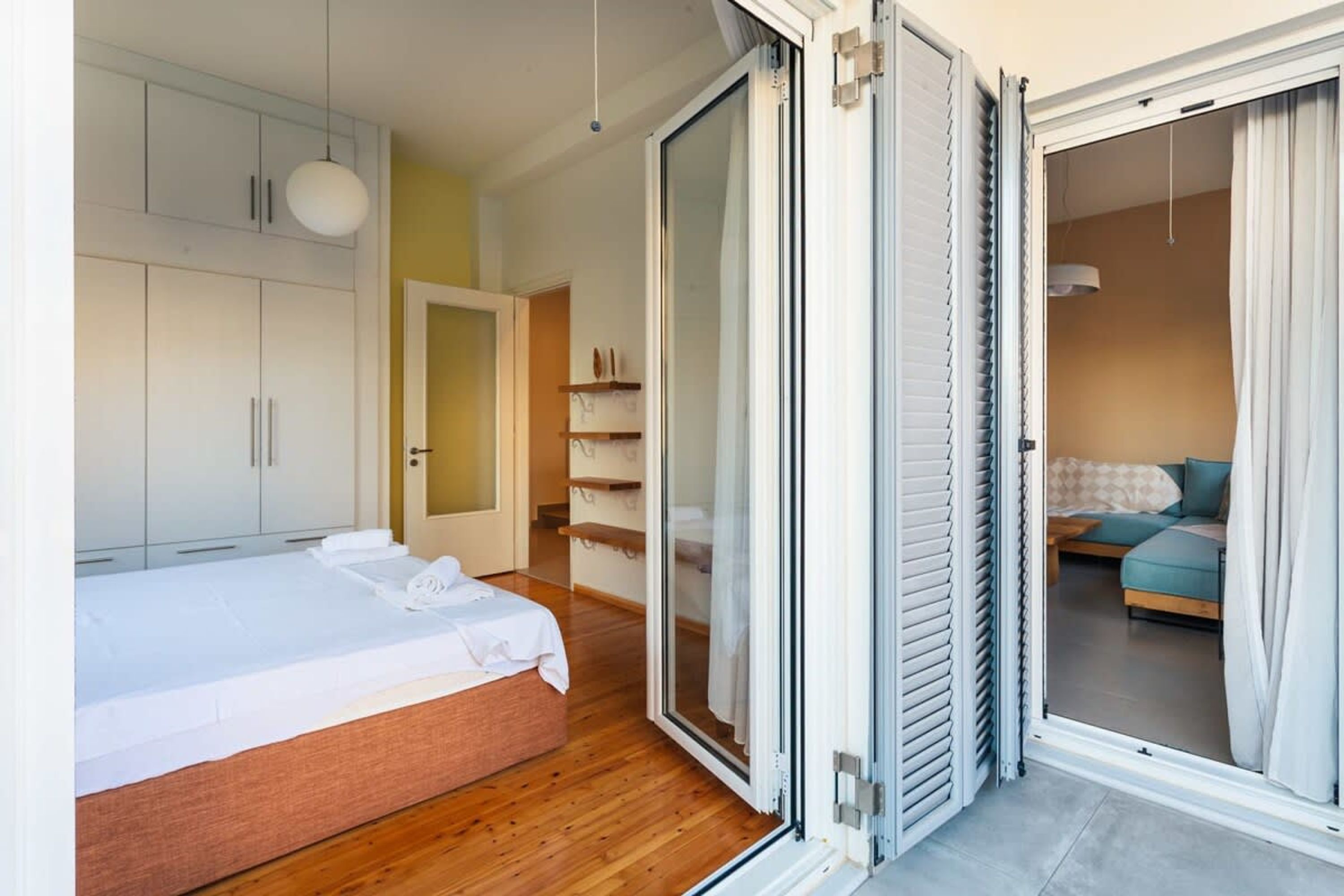 Quarto com cama, piso de madeira, guarda-roupa e porta de vidro deslizante para outro espaço.