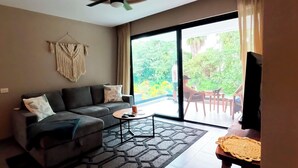 Uma sala de estar moderna com um sofá cinzento, uma mesa de centro e uma grande porta de vidro deslizante que dá para uma área externa com vegetação.