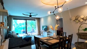 Uma sala de estar moderna com sofá, mesa de jantar e cadeiras. Há um ventilador de teto, uma televisão e uma janela grande com vista para o verde.