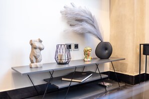 Um interior moderno com uma mesa consola cinzenta exibindo uma escultura de pedra, uma vela em suporte de arame, um busto decorativo e um vaso alto com uma pena.