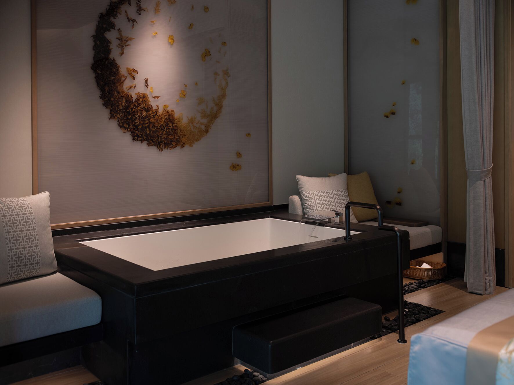 Uma casa de banho moderna com uma banheira grande, uma área de estar com almofadas e uma parede decorativa com folhas secas.