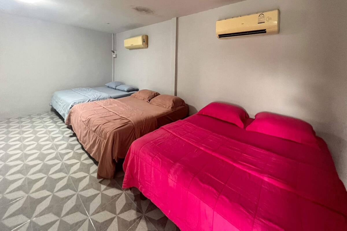 Quarto com duas camas, uma com colcha azul e outra com colcha vermelha, e dois aparelhos de ar condicionado na parede.