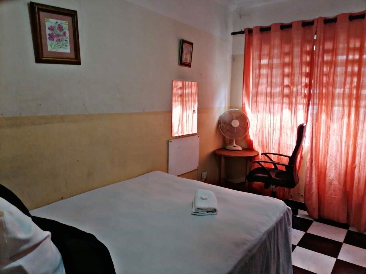 Quarto de hotel com uma cama, uma cadeira, uma mesa com um ventilador e uma janela com cortinas.