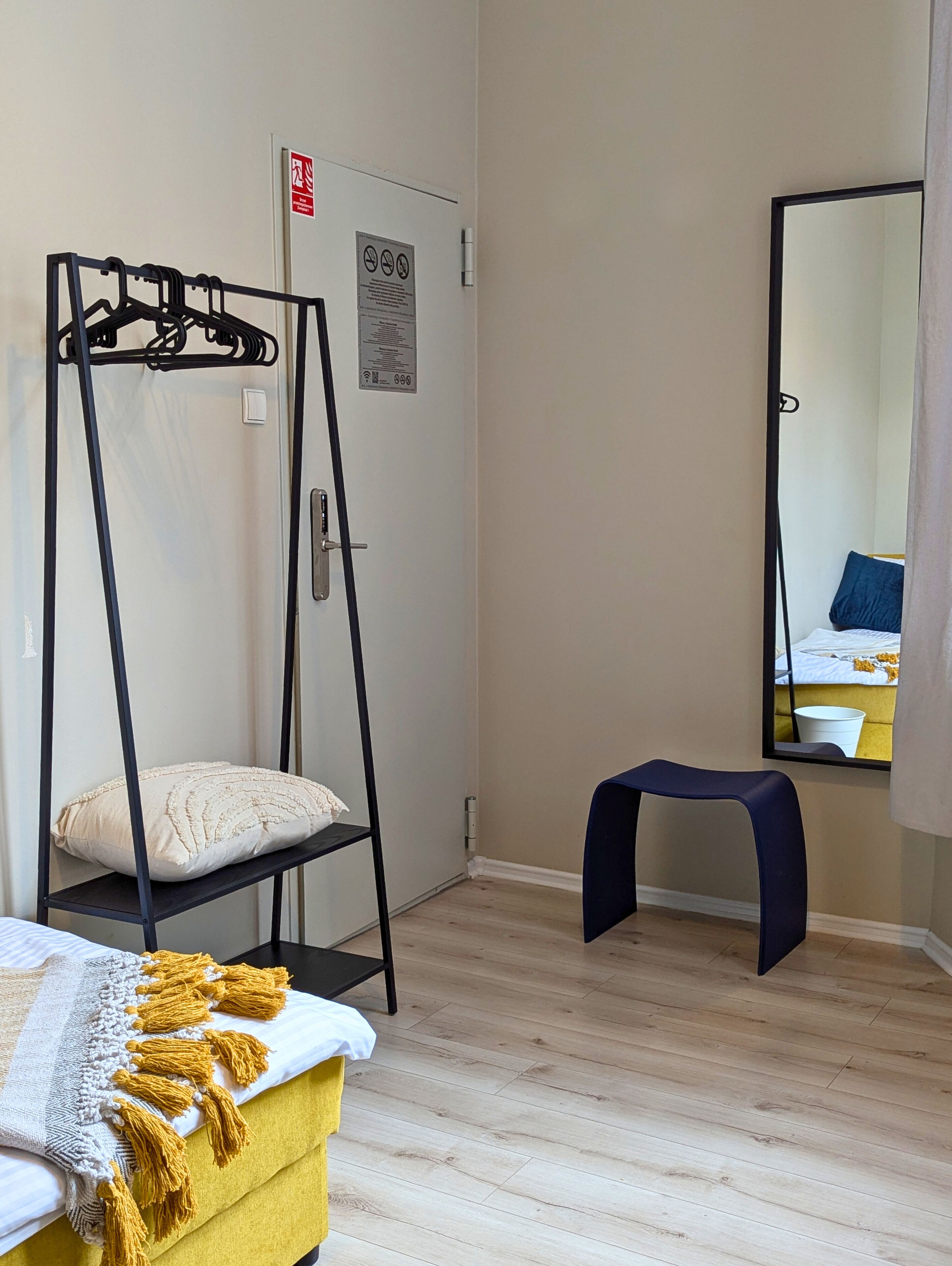 Quarto com cama beliche, um banquinho pequeno, um espelho e uma porta com sinal de segurança contra incêndio.