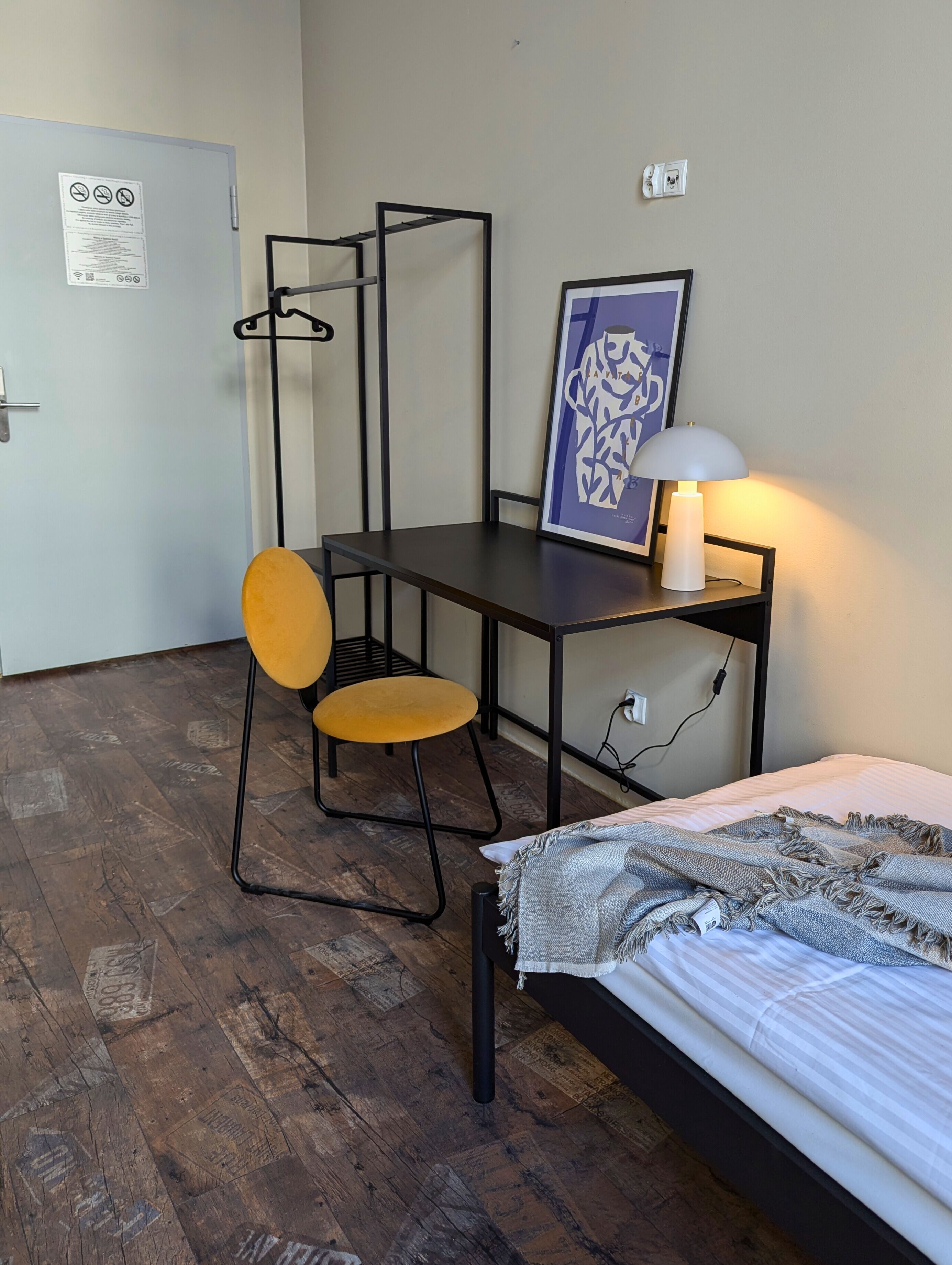 Quarto pequeno e bem organizado, com uma cama, uma escrivaninha com abajur, uma cadeira e um quadro na parede.