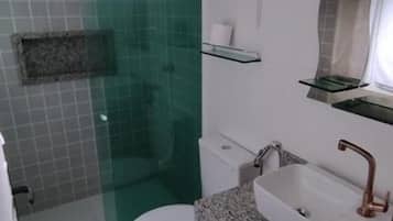 Uma casa de banho com cabine de duche em vidro, sanita, lavatório com espelho e prateleiras acima do lavatório.