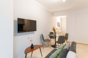 Uma sala de estar moderna com televisão de tela plana na parede, um espaço de jantar com mesa redonda e cadeiras, e um sofá com vários almofados.