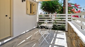 Um pátio com mesa e cadeiras brancas, uma pérgola com tecido amarelo e um cercado branco com plantas e flores.