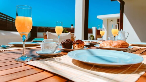 Uma mesa de café da manhã com copos de suco de laranja, uma xícara, muffins e croissants sobre uma mesa de madeira, com vista para o mar.