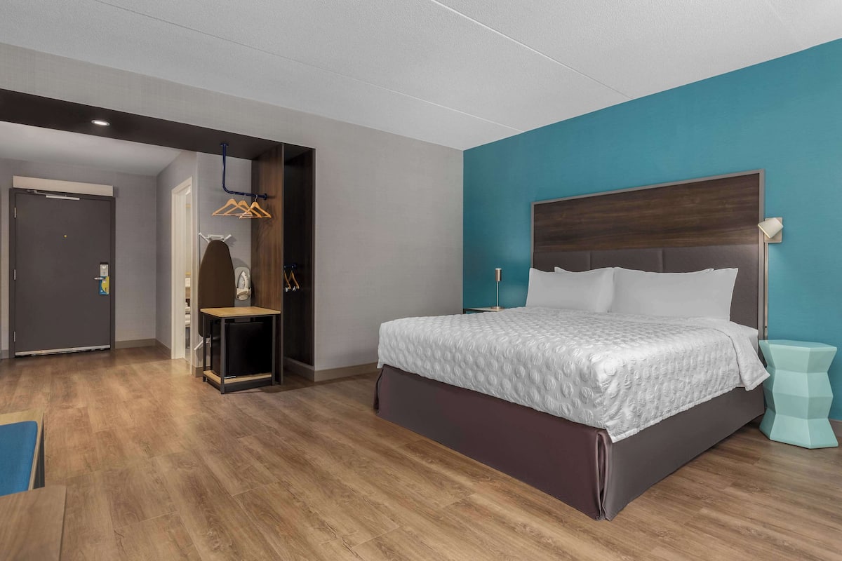Quarto de hotel moderno com cama grande, piso de madeira, parede em tom de verde-água e guarda-roupa com roupas penduradas.
