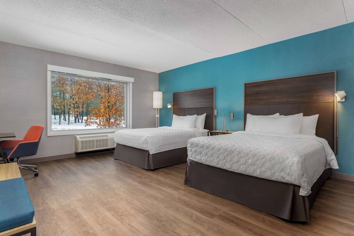 Quarto de hotel moderno com duas camas, uma escrivaninha e uma janela grande com vista para árvores de outono.