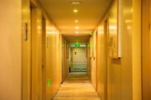 Corredor de hotel com portas em ambos os lados, piso coberto de carpete e sinalização de saída ao fundo.