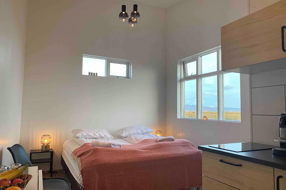 Quarto com cama, mesa de cabeceira, abajur e vista para o exterior pela janela.