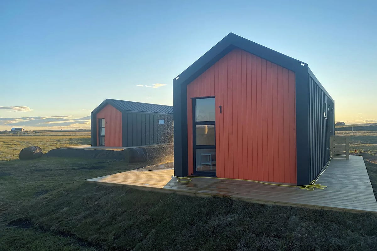 Duas cabanas modernas, em vermelho e preto, com decks de madeira, localizadas em um campo gramado sob um céu limpo.