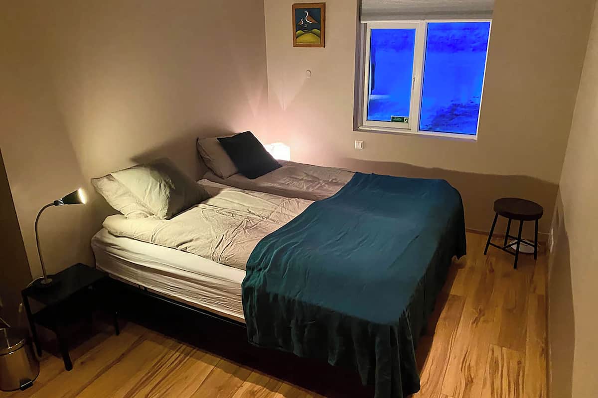 Uma cama de solteiro com um cobertor verde-água, uma luminária de cabeceira, um banquinho pequeno e uma janela com vista para o céu.