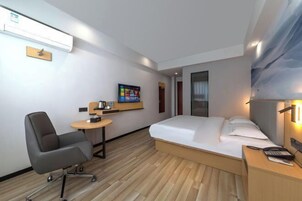 Quarto de hotel moderno com cama, escrivaninha, cadeira, mesa pequena e TV fixada na parede.