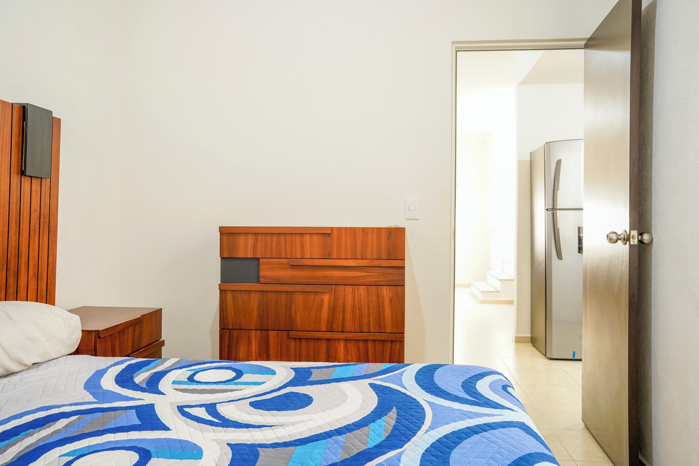 Quarto com cama, cómoda de madeira, roupeiro embutido e frigorífico moderno.