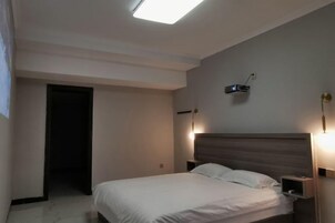 Quarto de hotel com cama, abajures, luminária no teto e porta que dá para outro cômodo.