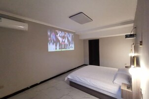 Quarto de hotel com uma cama, projetor fixado na parede e luminária.