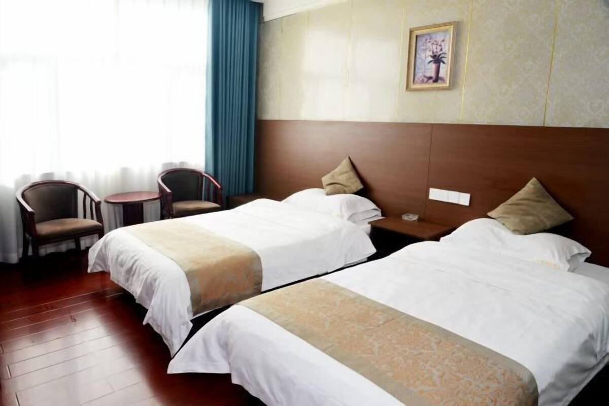 Quarto de hotel com duas camas, uma mesinha, uma cadeira e uma janela com cortinas.
