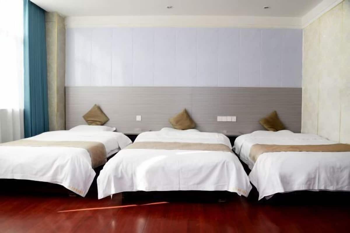 Quarto de hotel com três camas de solteiro, piso de madeira e uma grande janela com cortinas.