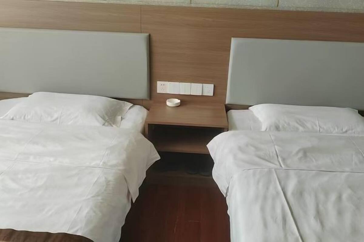 Quarto de hotel com duas camas de solteiro, cabeceira de madeira, mesa de cabeceira com abajur e uma pequena mesa.